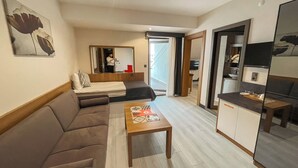 Quarto de hotel moderno com sofá, mesa de centro de madeira, cama, cozinha compacta e um espelho grande.