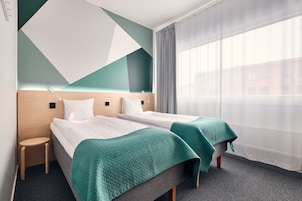 Quarto de hotel com duas camas, um banquinho pequeno e uma janela grande com cortinas.
