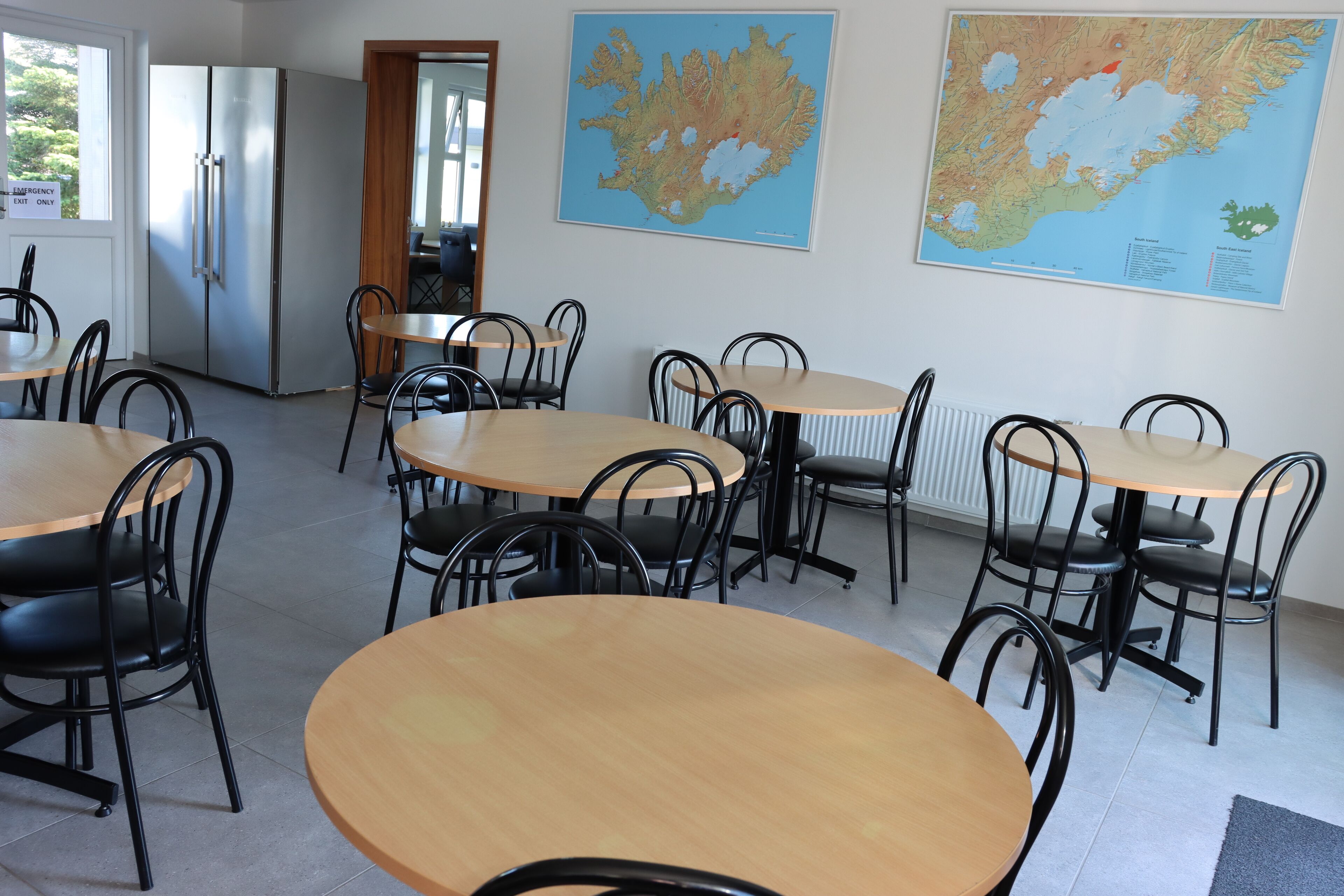Uma área de refeições com mesas redondas e cadeiras pretas, uma geladeira e dois mapas na parede.