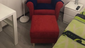 Quarto pequeno e bem organizado, com uma cadeira vermelha, uma mesa branca, um abajur, uma mesinha de cabeceira com luminária e uma cama com colcha estampada em verde e preto.