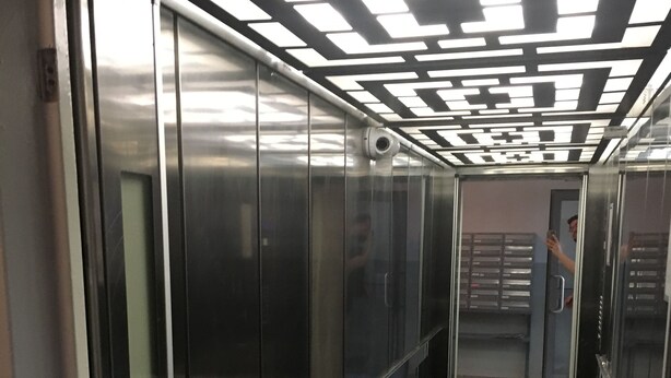 Interior de elevador moderno, com paredes metálicas, porta de vidro e teto estampado.