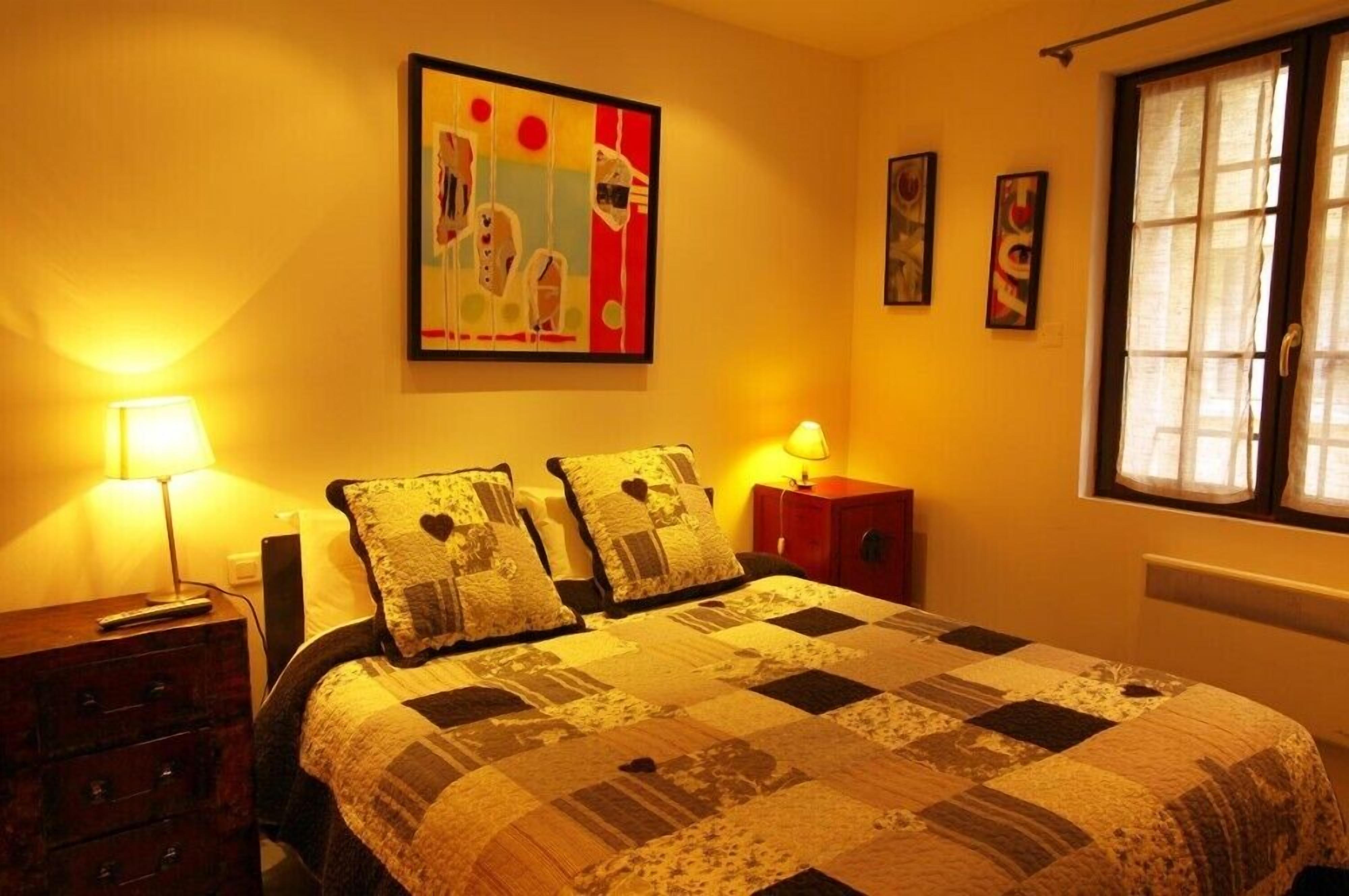 Quarto com edredom acolchoado, duas luminárias de cabeceira, cômoda e janela com cortinas.