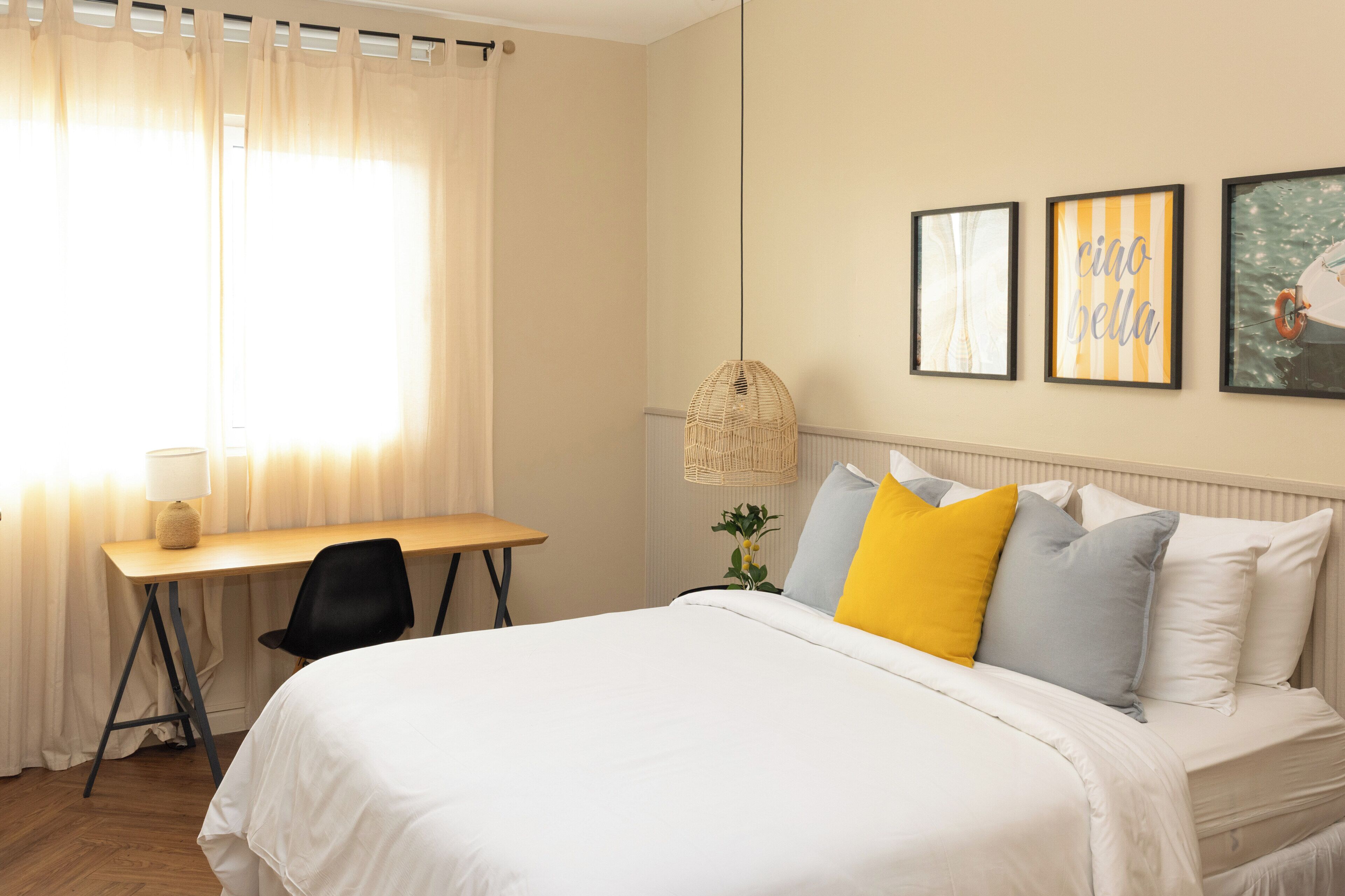 Quarto de hotel com uma cama, uma mesa, uma cadeira, um candeeiro pendente e duas imagens emolduradas na parede.