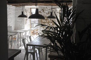 Uma área de jantar rústica com mesas de madeira e cadeiras brancas, grandes luminárias pendentes e um vaso com planta.