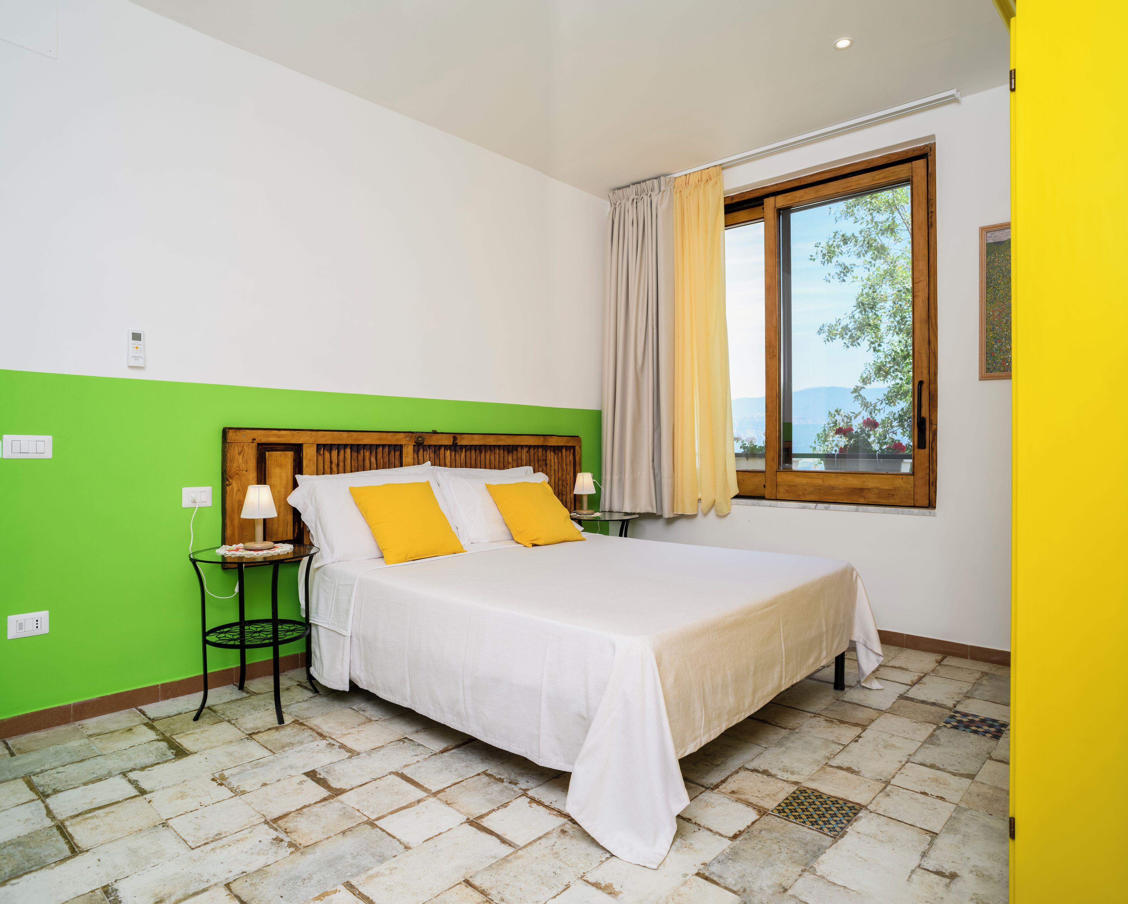 Quarto com cama, mesa de cabeceira, janela com cortinas e vista para o verde lá fora.