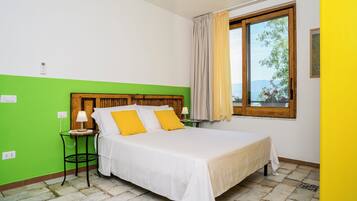 Quarto com cama, mesa de cabeceira, janela com cortinas e vista para o verde lá fora.