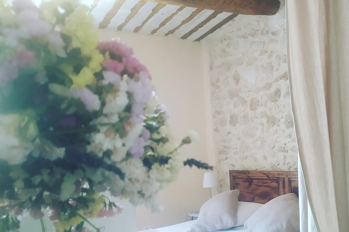 Quarto com cama, almofadas, mesa de cabeceira e um vaso com flores.
