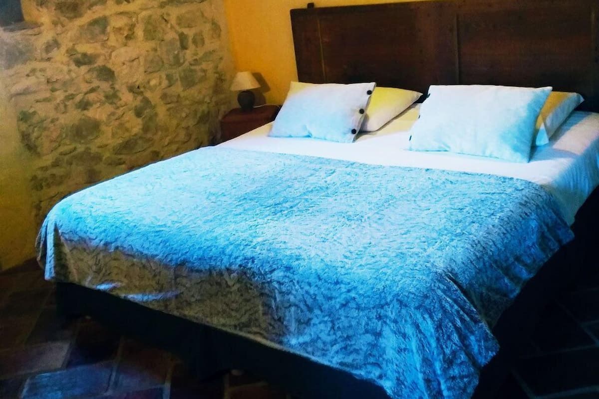 Uma cama com colcha estampada em azul, cabeceira de madeira e mesa de cabeceira com uma luminária. O quarto tem paredes de pedra e piso ladrilhado.