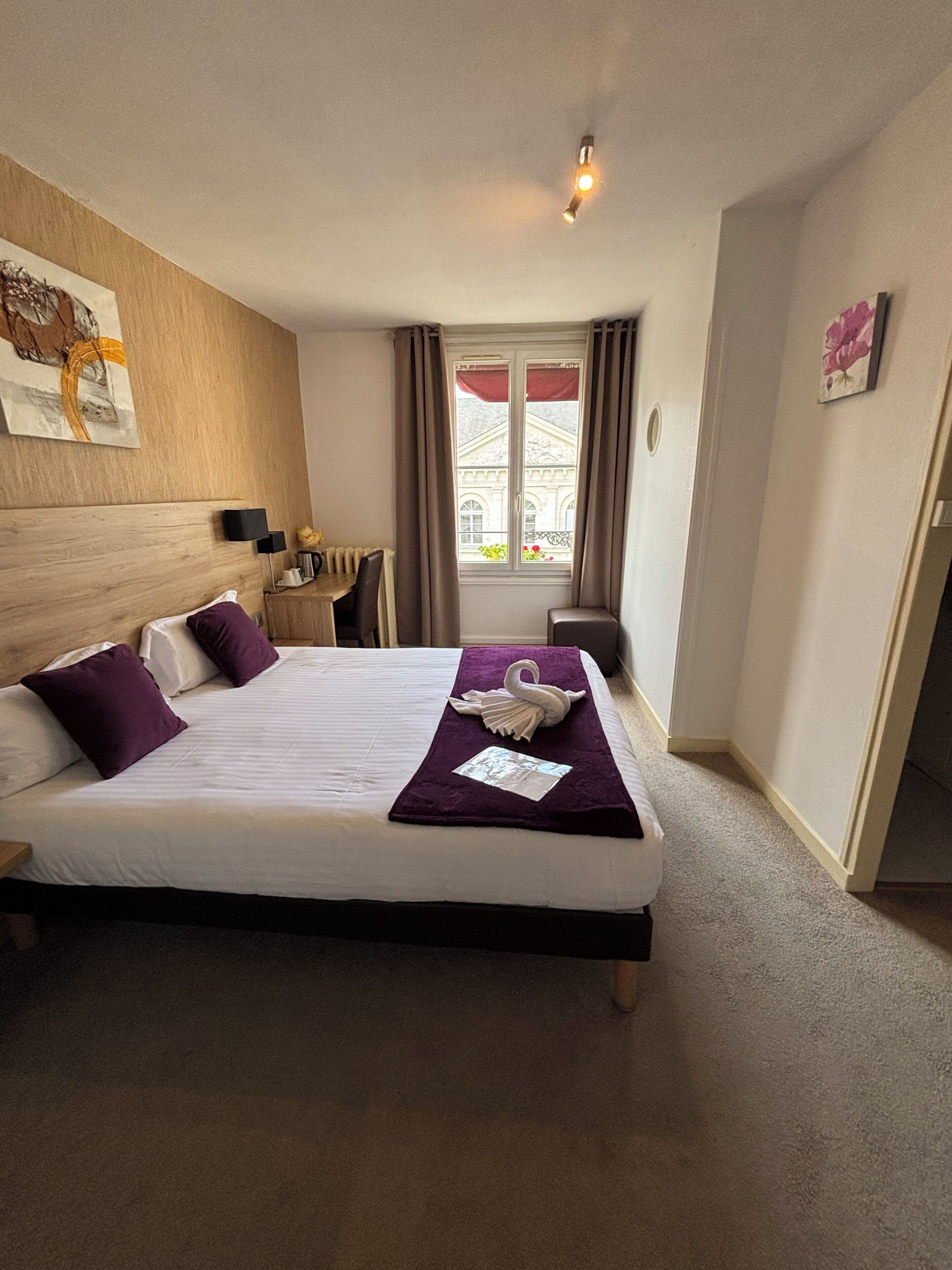 Quarto de hotel com uma cama, uma escrivaninha, uma cadeira, janela com cortinas e um quadro na parede.