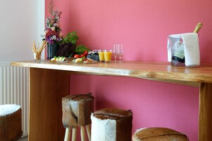 Uma mesa de madeira com uma parede rosa ao fundo, um vaso de flores, dois copos de suco de laranja e uma garrafa de champanhe.