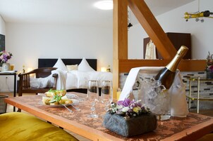 Quarto de hotel com cama, mesa de jantar, garrafa de champanhe, copos e um arranjo floral.
