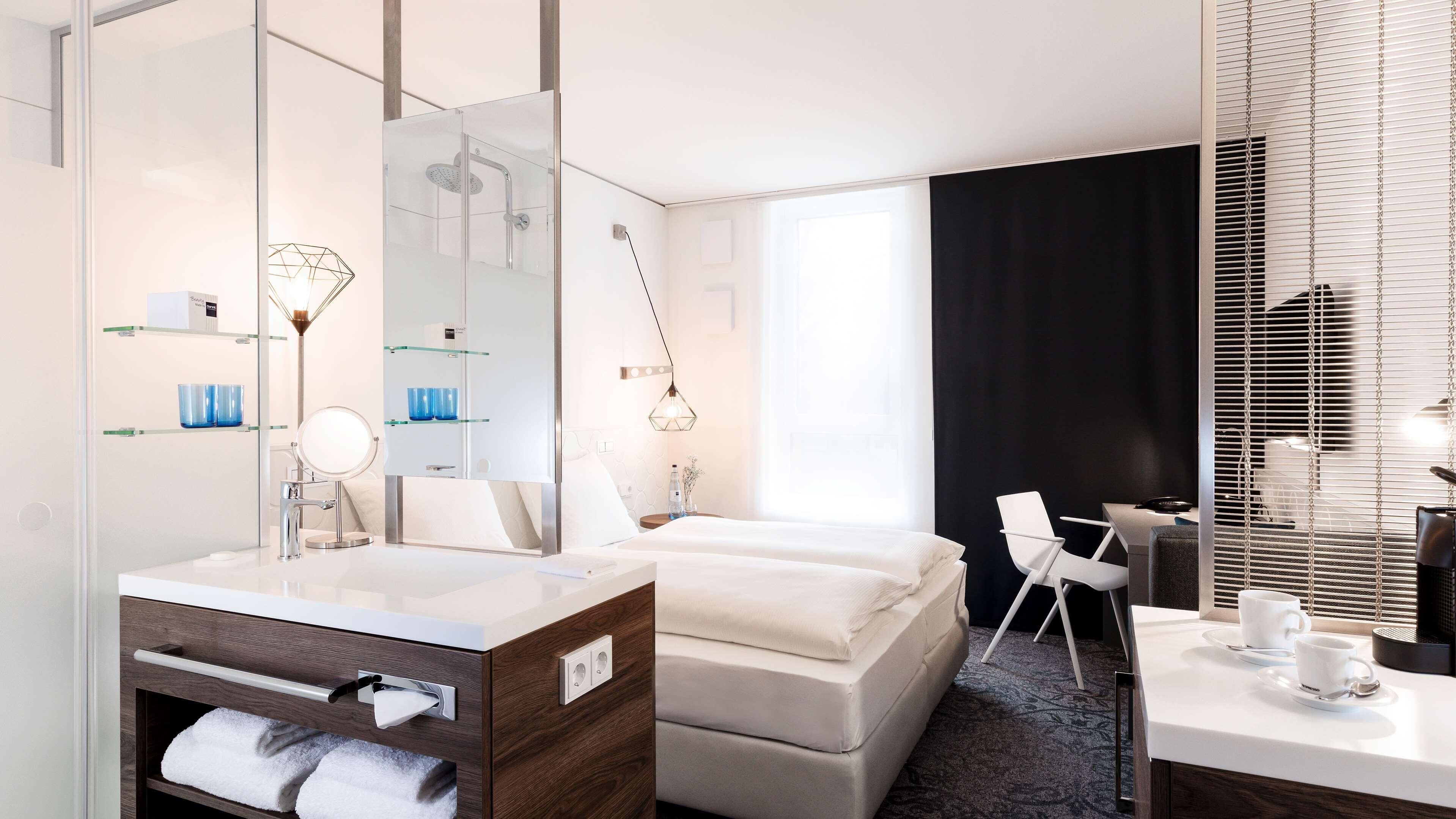 Quarto de hotel com uma cama de casal, uma escrivaninha, uma cadeira, um espelho grande e banheiro com pia e chuveiro.