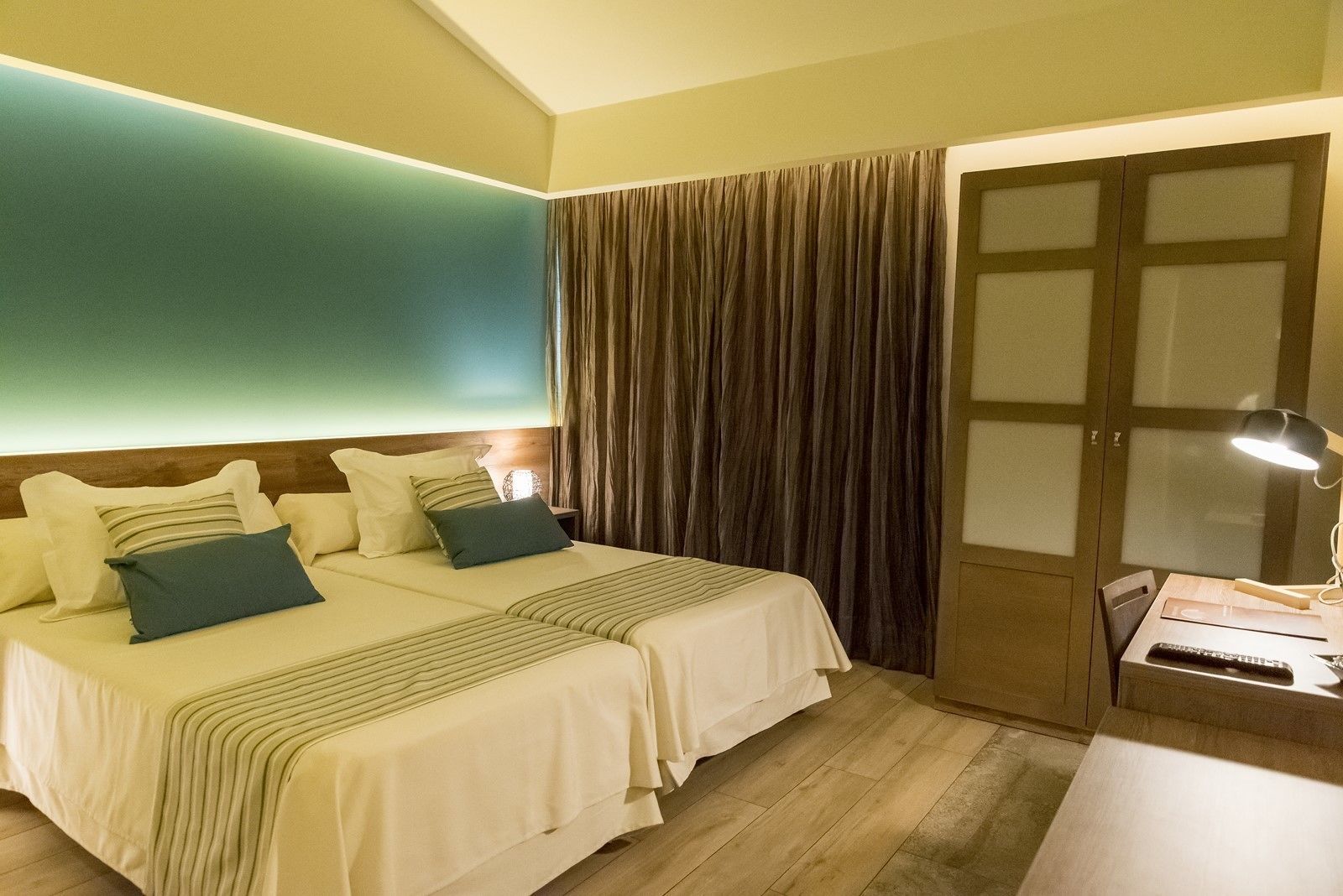 Quarto de hotel com duas camas, uma escrivaninha, uma luminária e uma cortina.