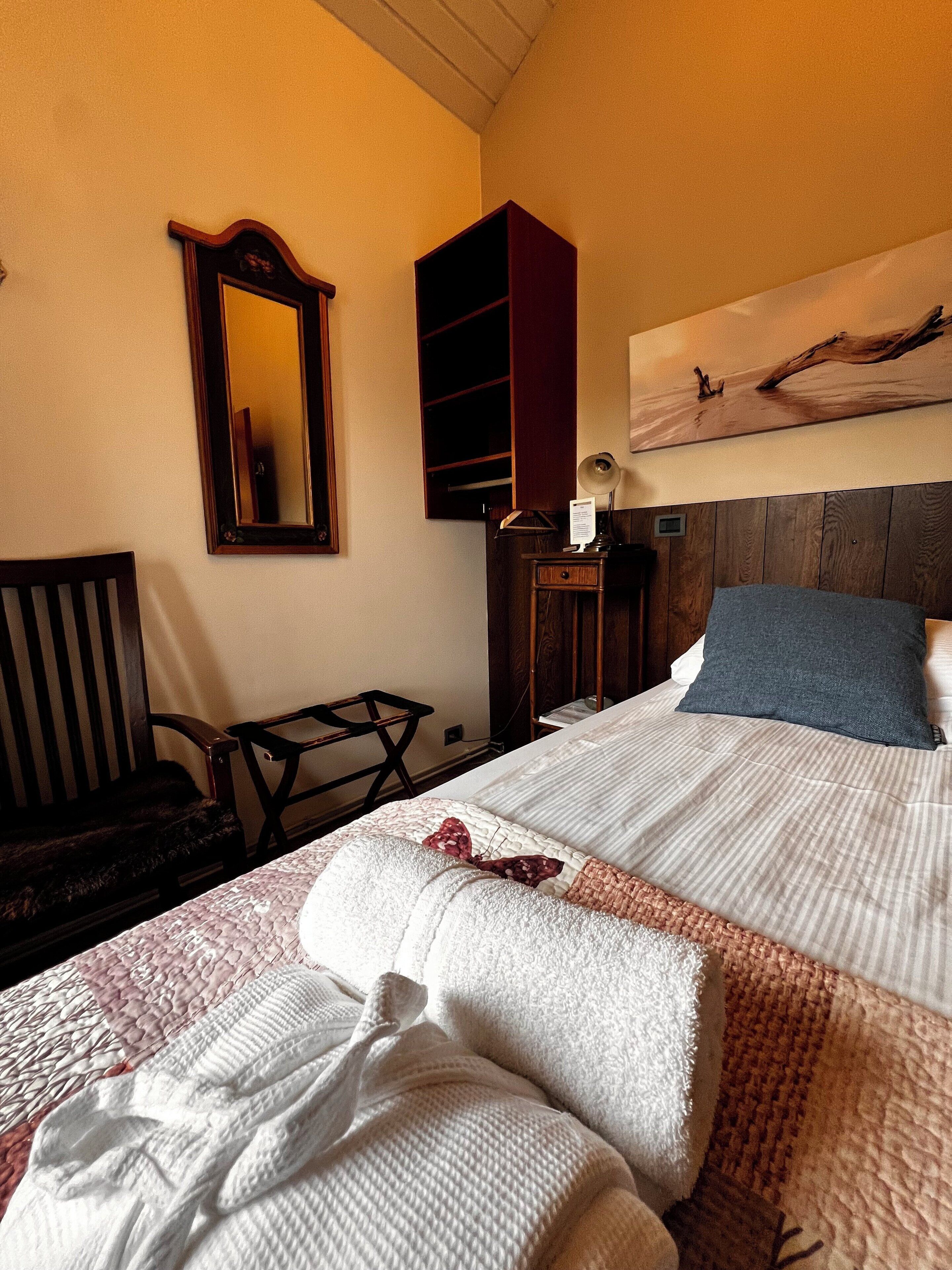 Quarto de hotel com uma cama grande, cabeceira de madeira, escrivaninha com cadeira, espelho e um quadro na parede.
