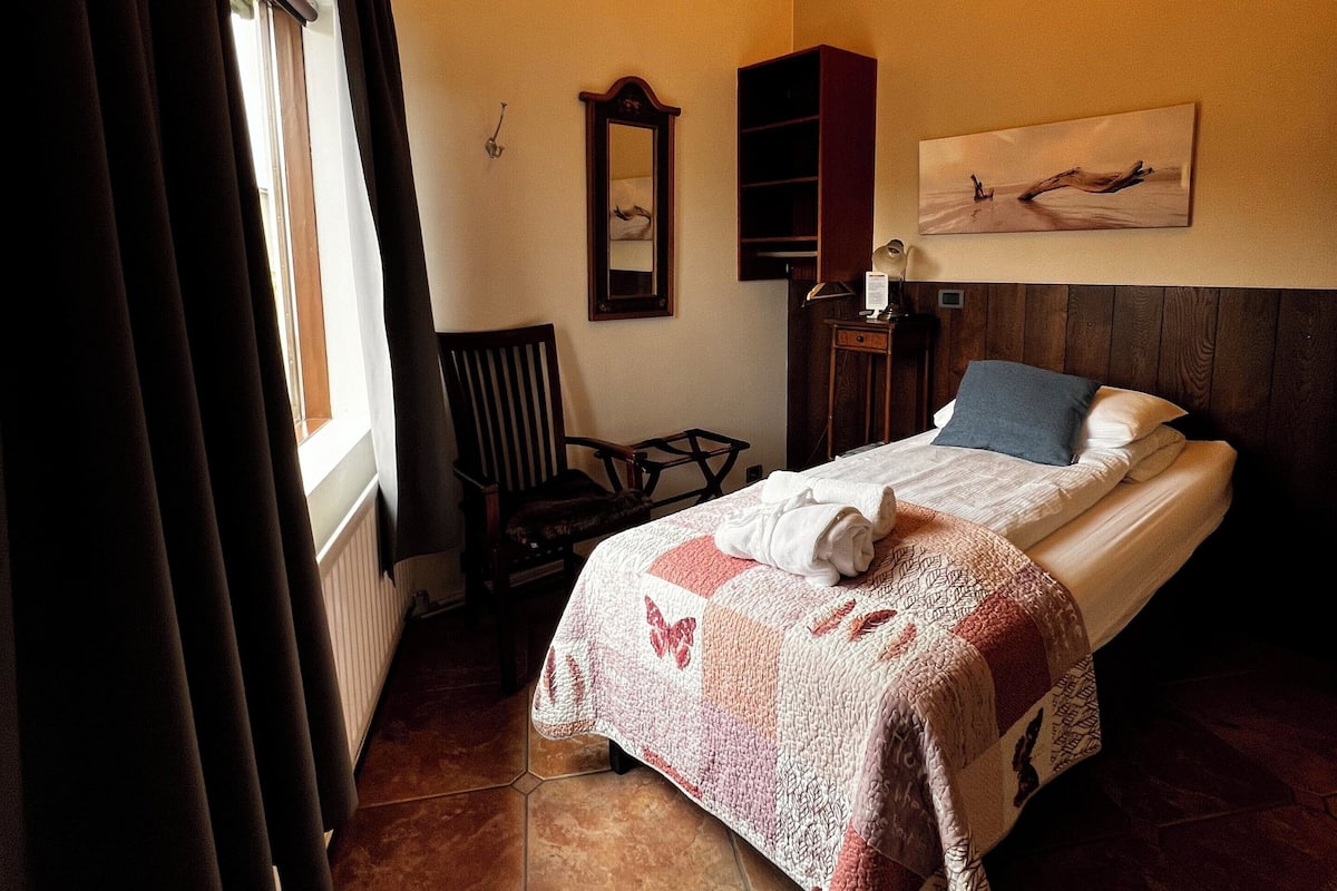 Quarto com cama, uma cadeira, um armário de madeira, um quadro e um candelabro.