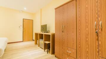 Quarto de hotel com piso de madeira, uma cama, um guarda-roupa, uma escrivaninha com computador e uma pequena mesa.