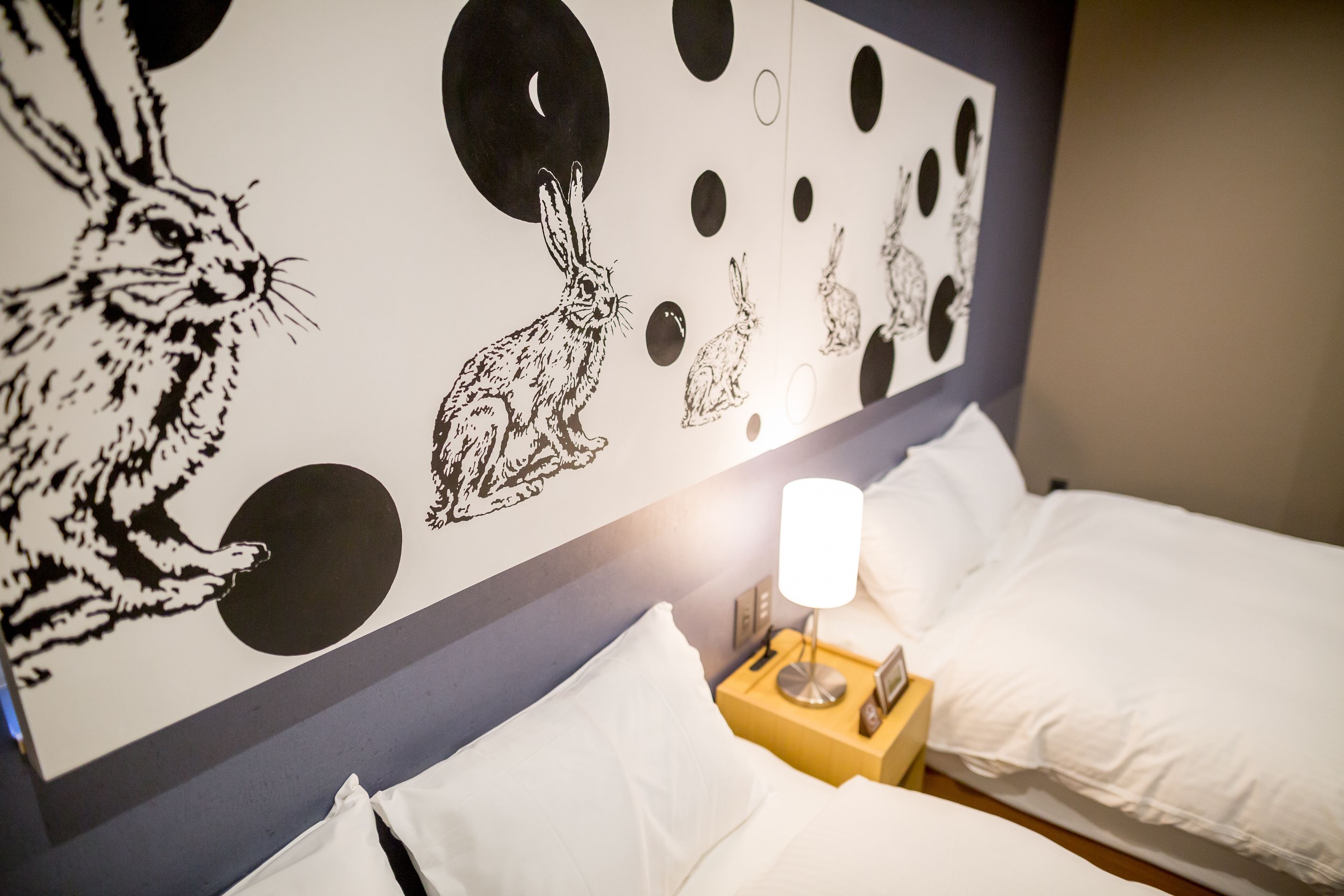 Quarto de hotel com um grande mural de coelhos na parede, uma mesinha de cabeceira com abajur e roupa de cama branca.