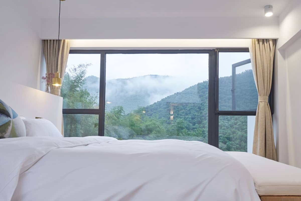 Quarto com cama, janela com cortinas e vista para uma paisagem montanhosa.