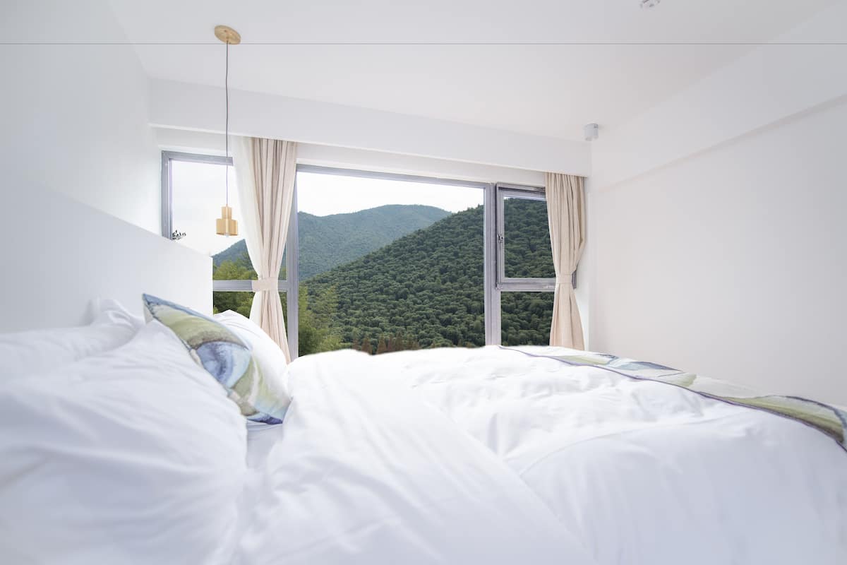 Quarto com cama, janela e vista para uma montanha arborizada.