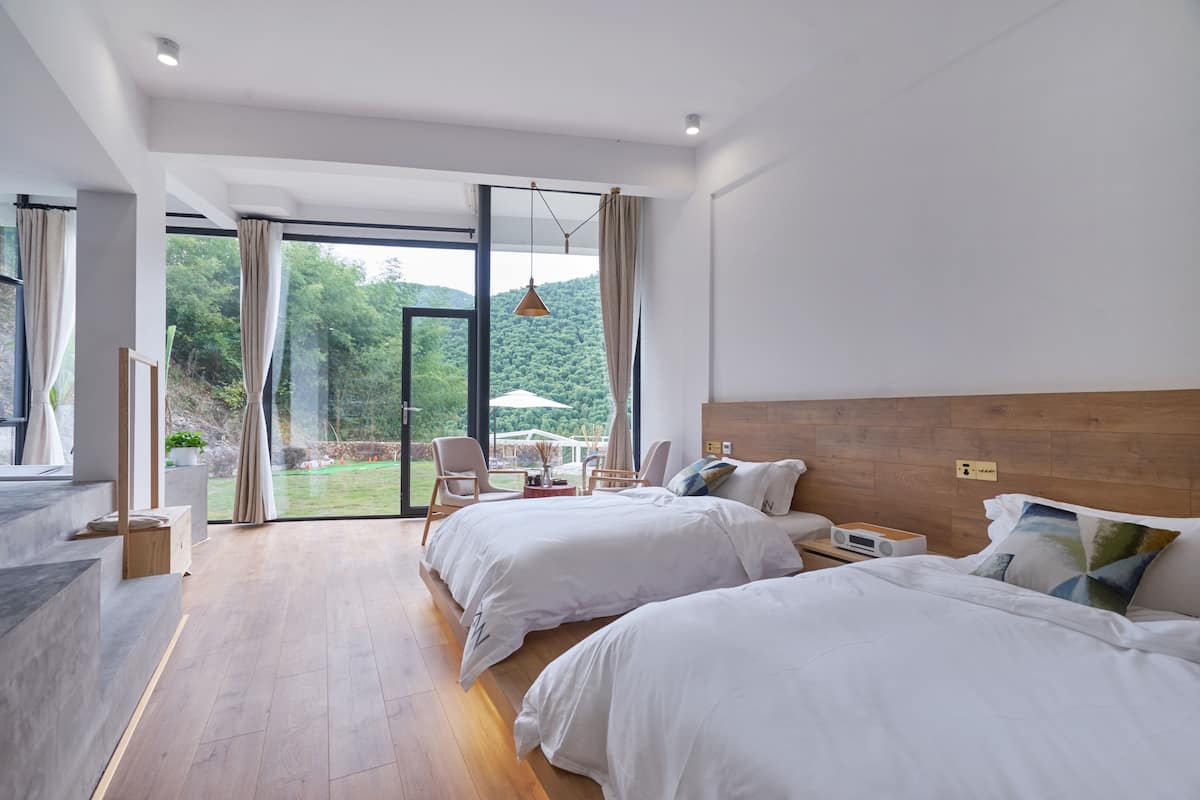 Quarto moderno com duas camas, cabeceira de madeira, uma janela grande com vista para vegetação e uma porta de vidro deslizante que dá para uma área externa.