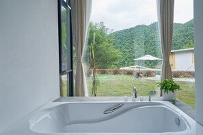 Um banheiro com uma banheira grande, janela com cortinas e vista para uma paisagem verde e um edifício ao fundo.