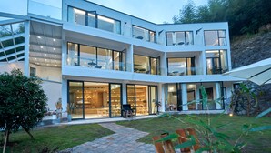 Uma casa moderna de vários andares com amplas janelas de vidro, área de estar externa e um jardim bem cuidado.