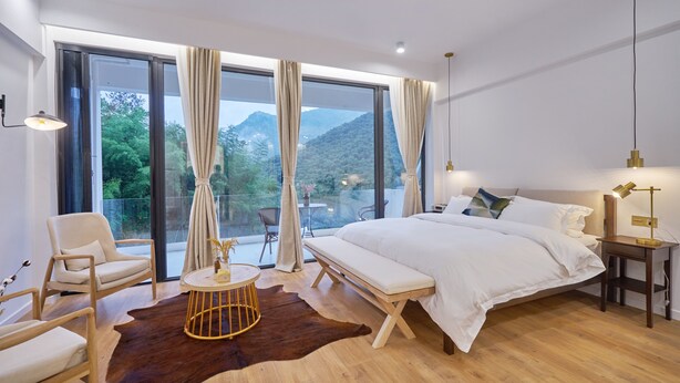 Um quarto moderno com uma cama grande, um banco de madeira, uma mesa redonda e vista para uma paisagem montanhosa através de amplas janelas.