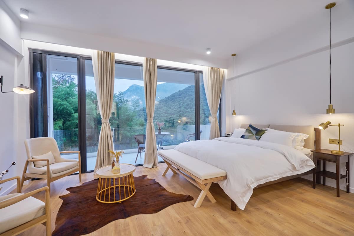 Um quarto moderno com uma cama grande, um banco de madeira, uma mesa redonda e vista para uma paisagem montanhosa através de amplas janelas.