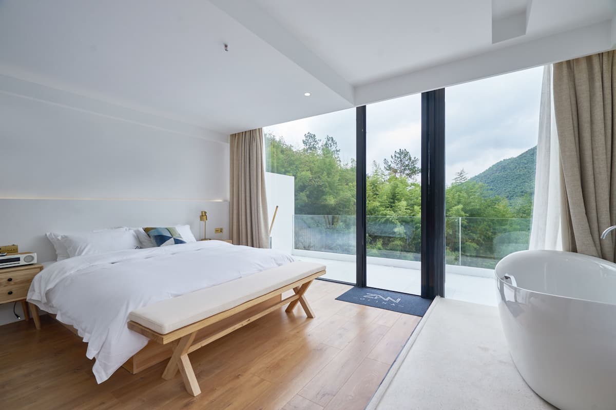 Quarto moderno com uma cama grande, um banquinho, uma banheira e vista para o verde através de janelas amplas.