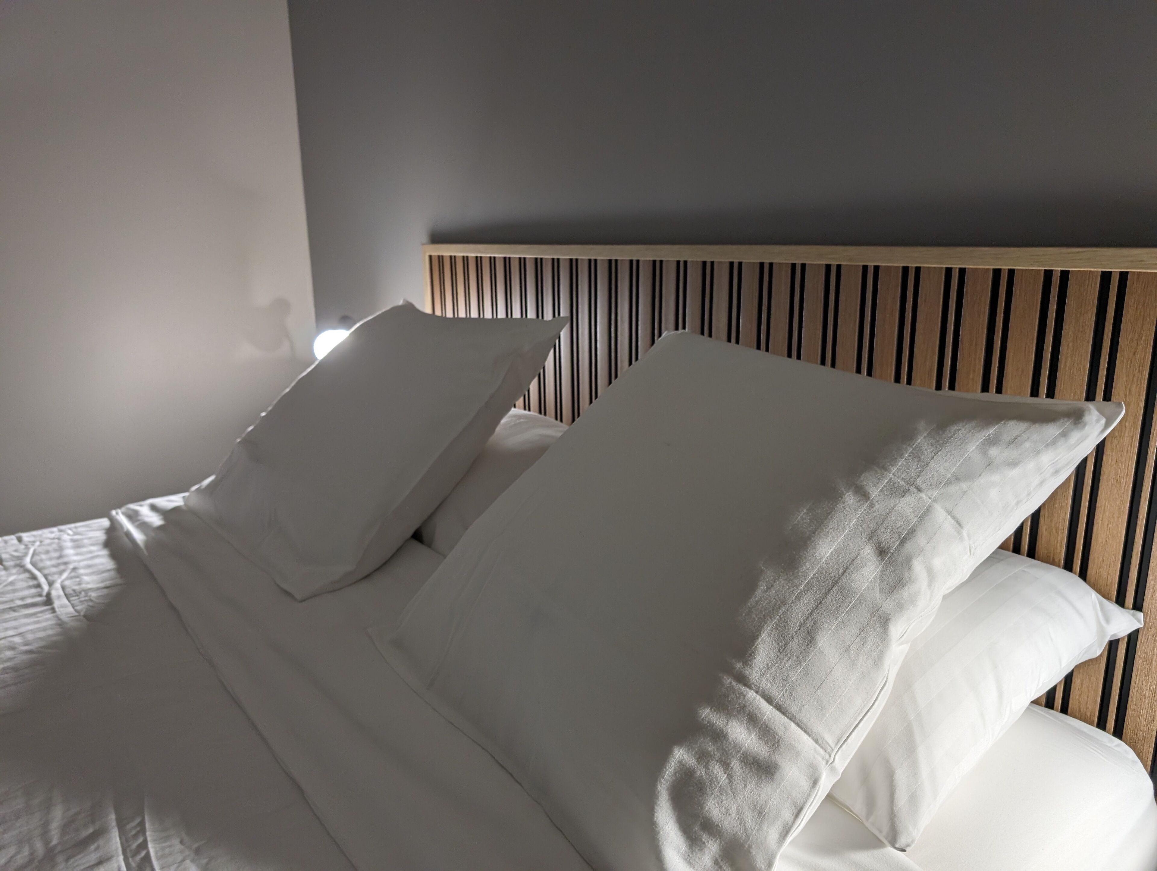Cama bem arrumada com lençóis e almofadas brancas, cabeceira de ripas de madeira e parede lisa.