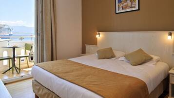Quarto de hotel com uma cama, duas almofadas, uma mesinha de cabeceira, um abajur, uma janela com cortinas, vista para um navio de cruzeiro e uma pequena varanda com mesa e cadeiras.