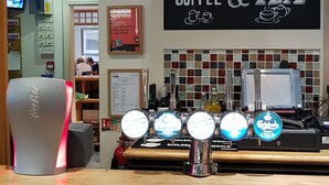 Interior de uma loja de café e chá, com um balcão exibindo diversas opções de bebidas, uma placa com a inscrição 'Yorkshire Coffee & Tea' e um refrigerador com a marca Coca-Cola.