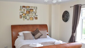 Cama bem arrumada com cabeceira marrom, roupa de cama branca e duas almofadas. Um objeto decorativo está acima da cama, e uma cortina está parcialmente aberta.