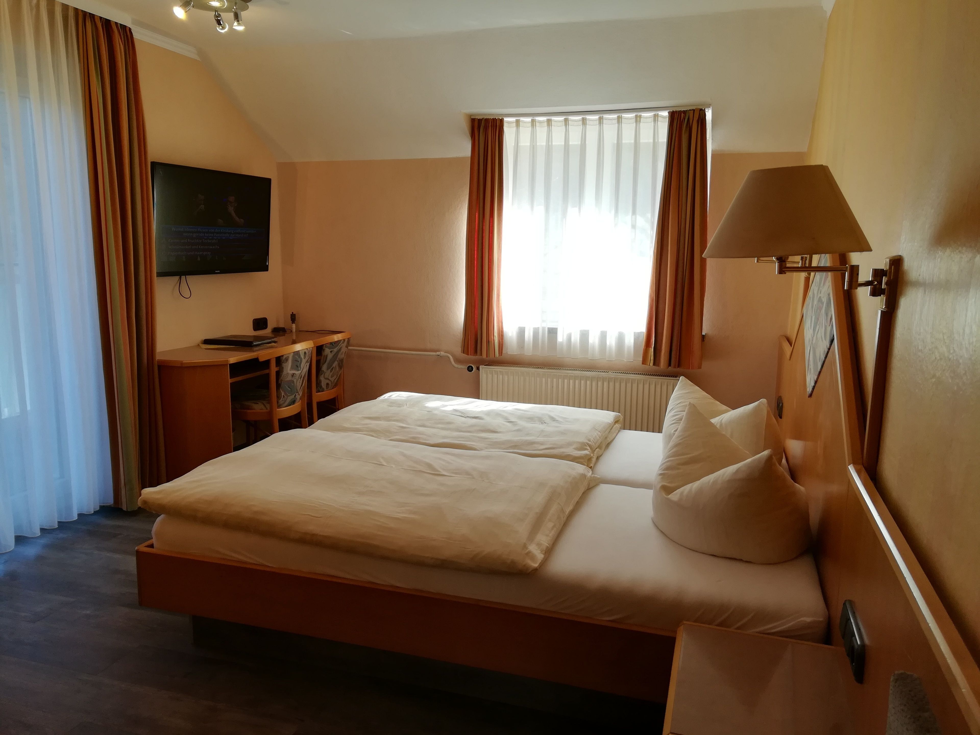 Quarto de hotel com cama de casal, escrivaninha com cadeira, televisão e janela com cortinas.