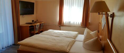 Quarto de hotel com cama de casal, escrivaninha com cadeira, televisão e janela com cortinas.
