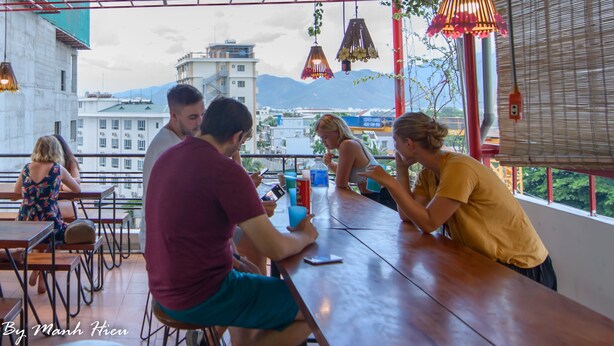 Um grupo de pessoas sentadas em uma mesa de madeira numa área de refeições ao ar livre, com vista para a cidade, sob luminárias pendentes.