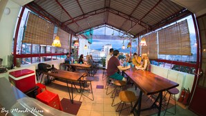 Um restaurante com mesas e cadeiras, janelas amplas e luminárias pendentes.