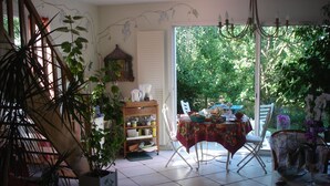 Quarto iluminado pelo sol com uma mesa de jantar posta para o café da manhã, rodeado por plantas em vasos e uma grande janela com vista para o verde.