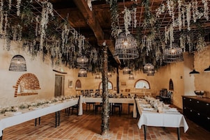 Uma longa mesa de jantar posta com toalhas brancas e cadeiras, rodeada por decorações florais pendentes e paredes de tijolos rústicos.