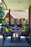 Um pátio externo com uma mesa posta para dois, rodeado por cadeiras de vime, uma área coberta e um edifício vermelho ao fundo.