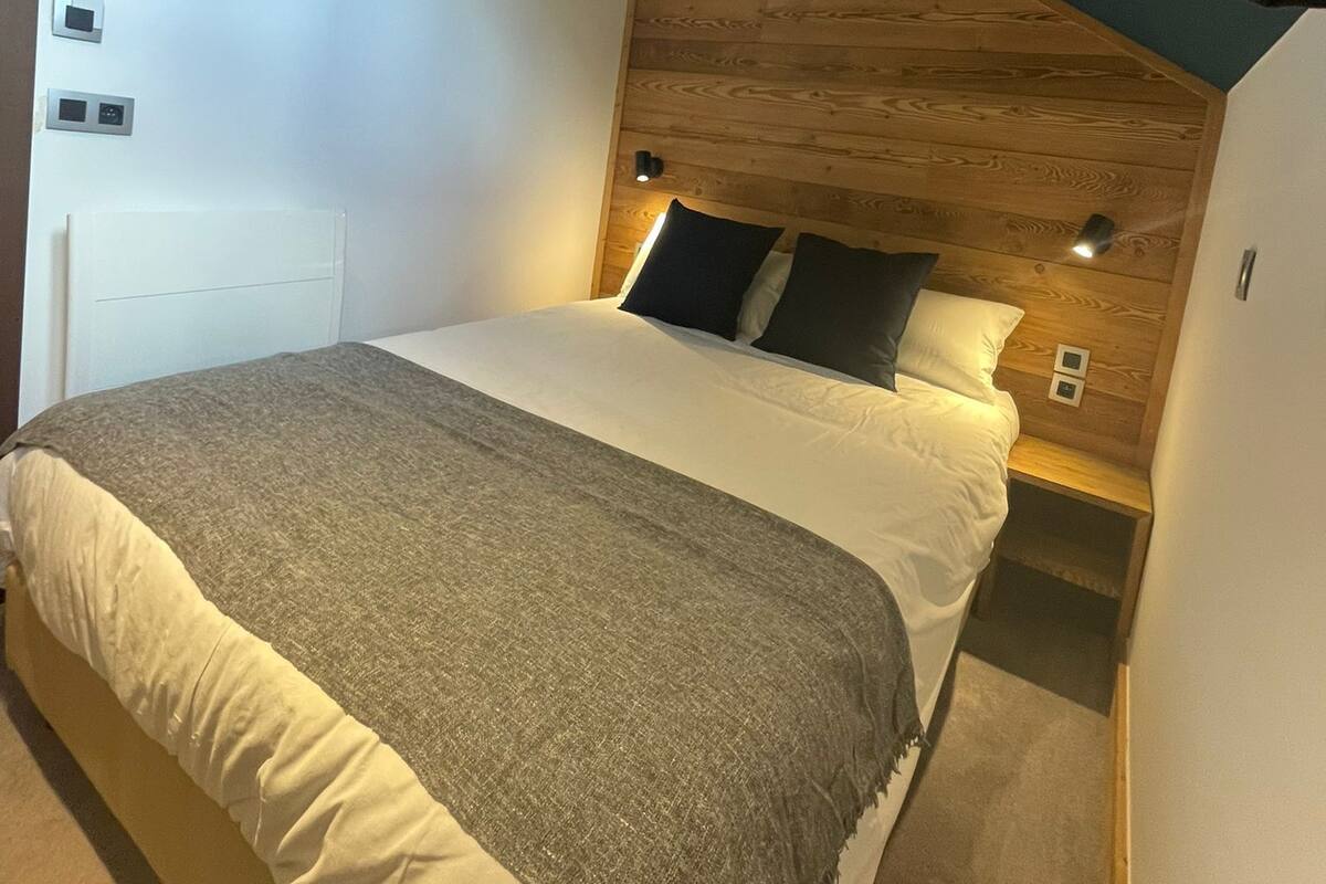 Quarto com cama, cabeceira de madeira e parede branca com termostato.