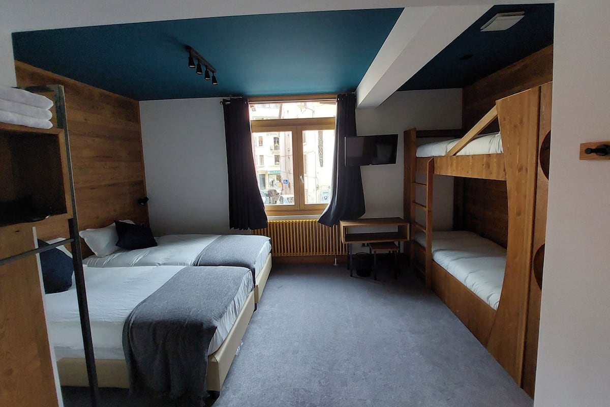 Quarto pequeno com beliches, escrivaninha e janela com vista para edifícios.
