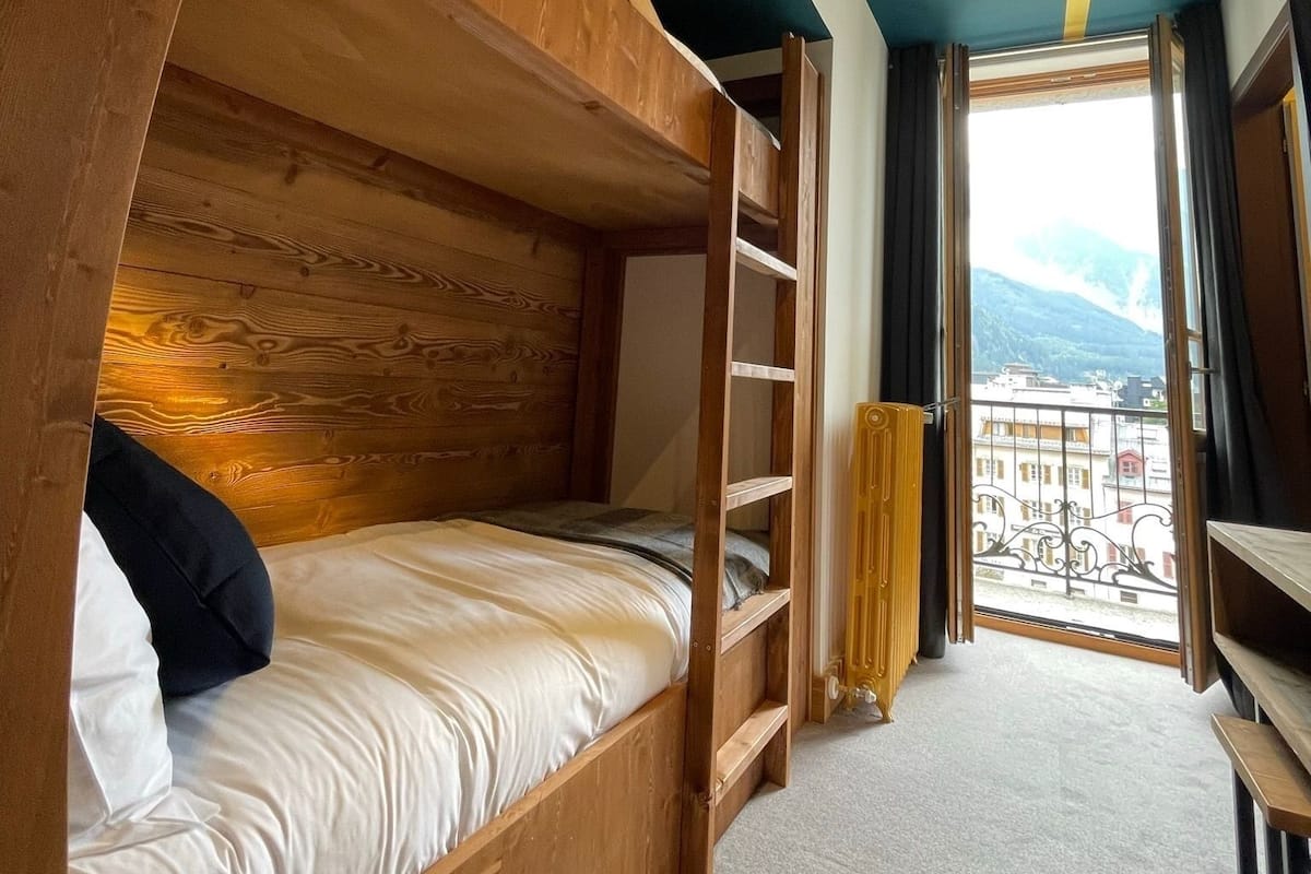 Quarto com beliche, escrivaninha de madeira, cadeira, radiador e vista para edifícios pela janela.