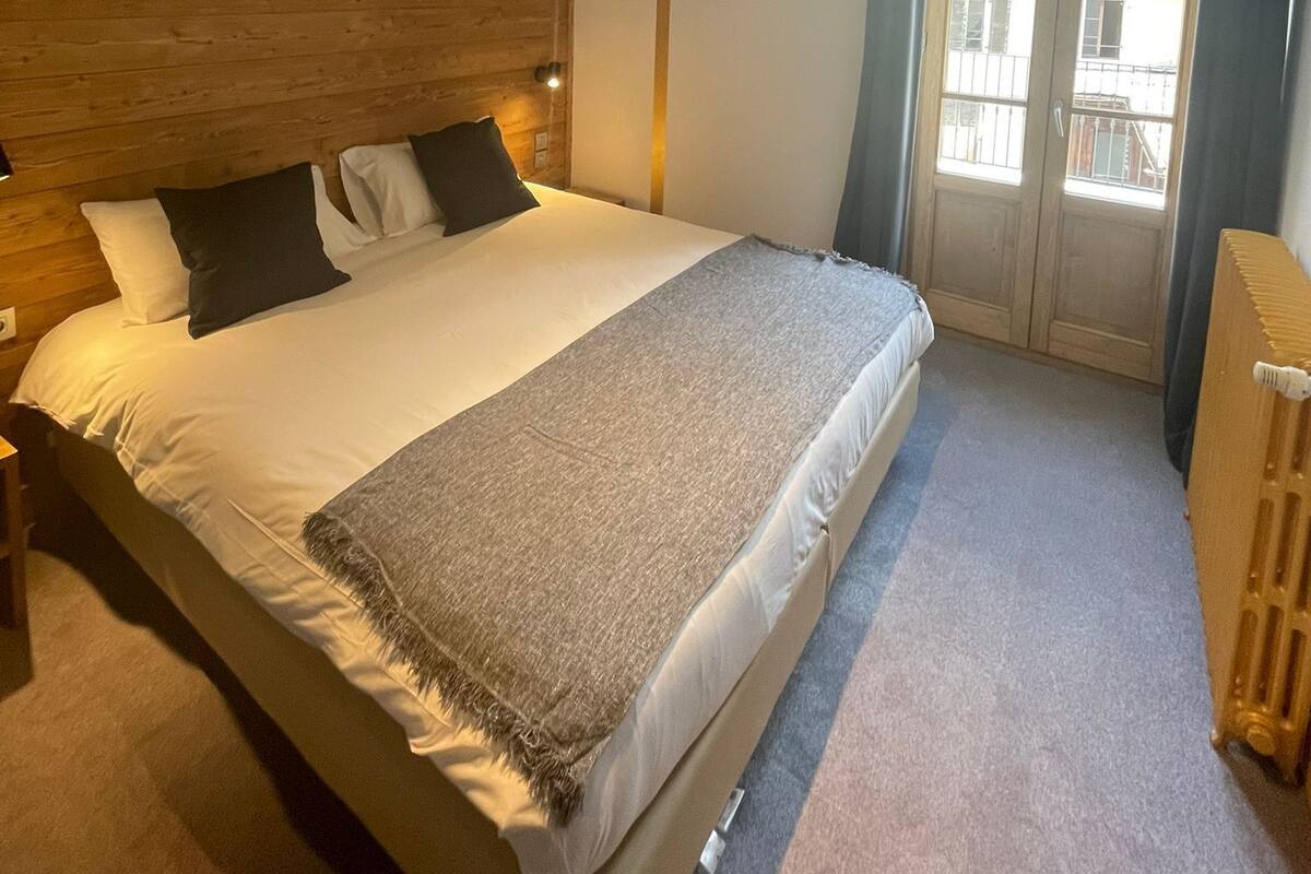 Quarto com cabeceira de madeira, uma cama grande com roupa de cama branca e um cobertor cinza, mesa de cabeceira de madeira e janela com vista para prédios.