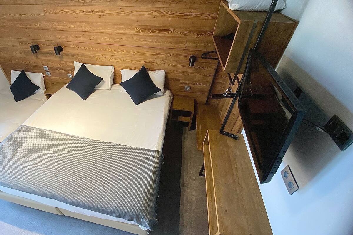 Quarto pequeno e compacto com uma cama de solteiro, cabeceira de madeira e televisão fixada na parede.