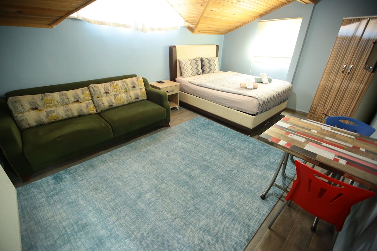Um quarto pequeno e bem iluminado, com um sofá verde, uma cama com colcha estampada, um guarda-roupa de madeira e uma pequena mesa com uma cadeira.