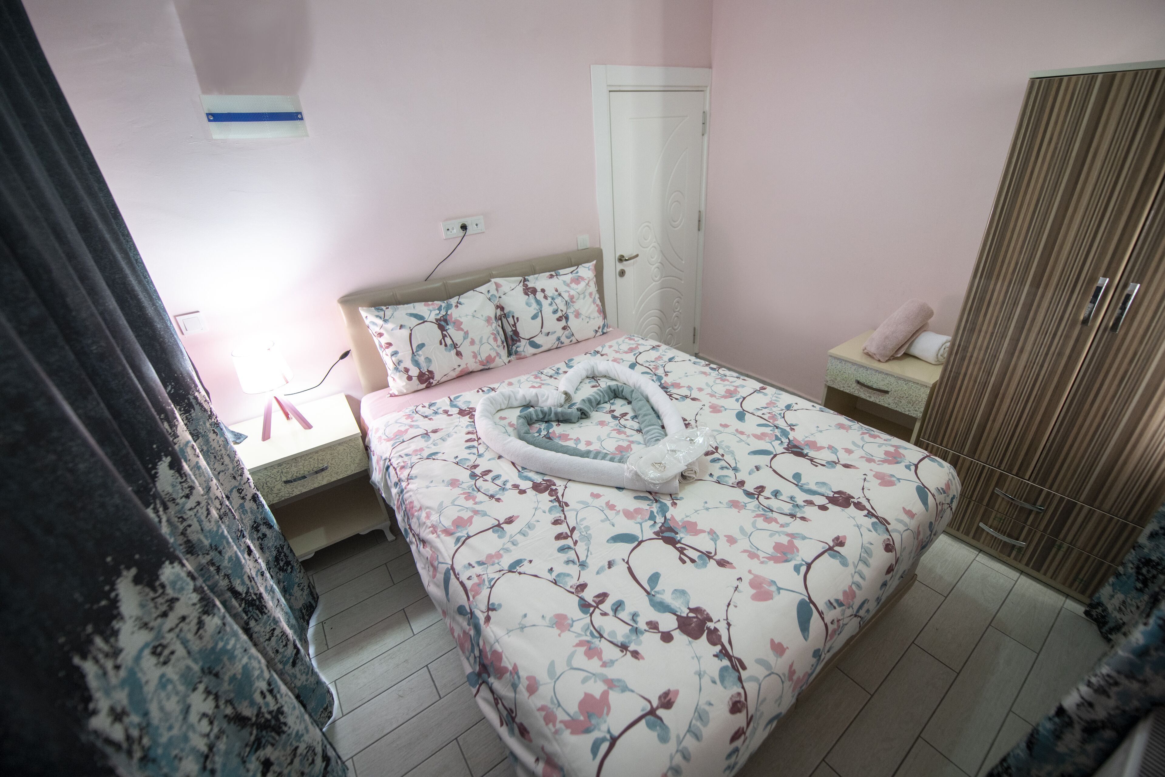 Quarto com colcha florida, mesa de cabeceira com abajur, guarda-roupa e porta com puxador decorativo.