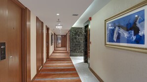Corredor de hotel com portas de madeira, carpete listrado, parede de mármore e uma obra de arte emoldurada.