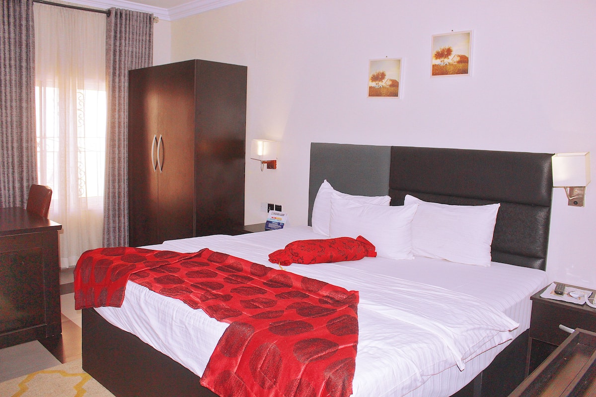 Quarto de hotel com uma cama grande, um guarda-roupa, uma mesa de cabeceira com uma luminária e duas fotografias emolduradas na parede.