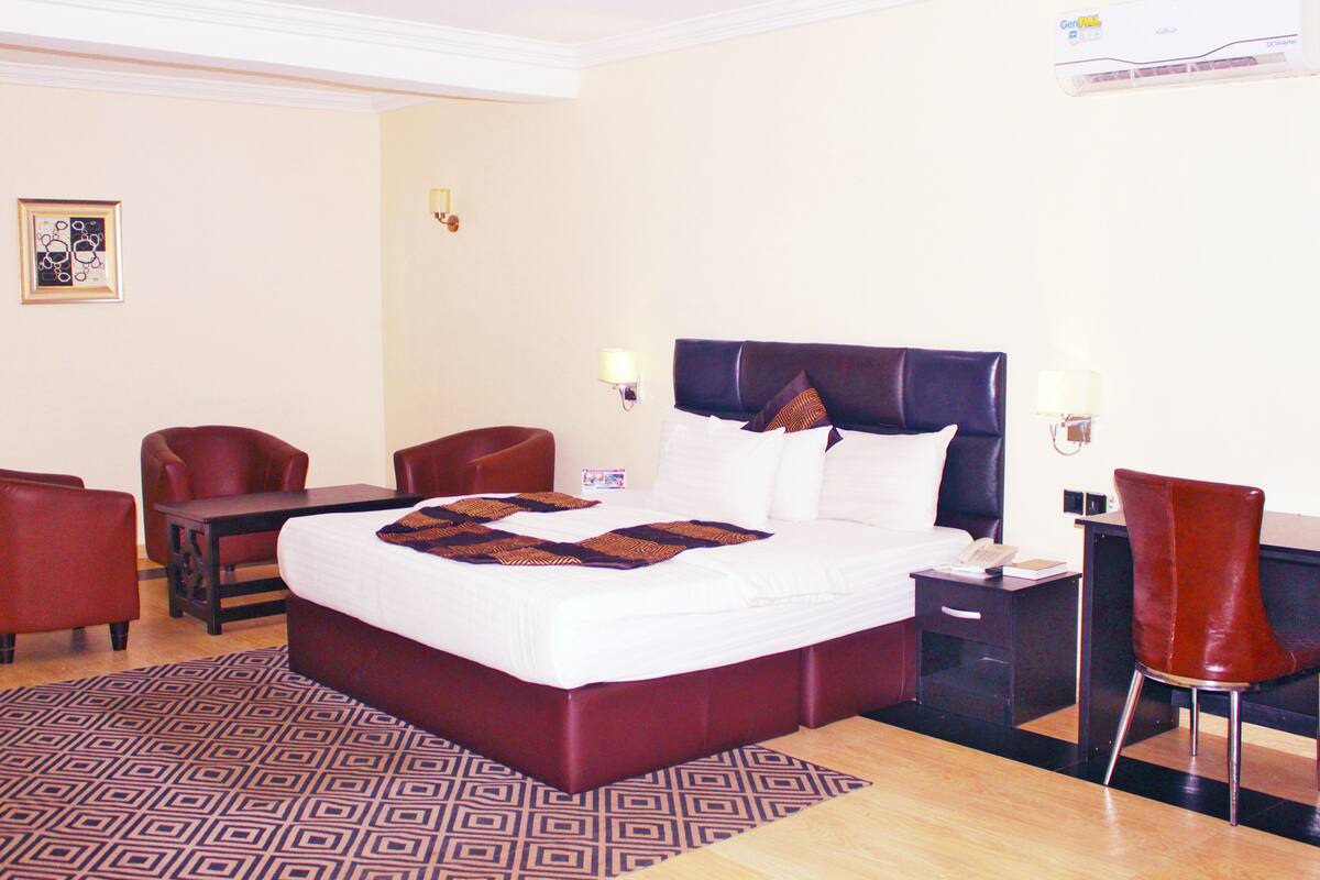 Quarto de hotel com uma cama grande, uma escrivaninha, duas cadeiras, uma mesa de cabeceira e um tapete estampado.